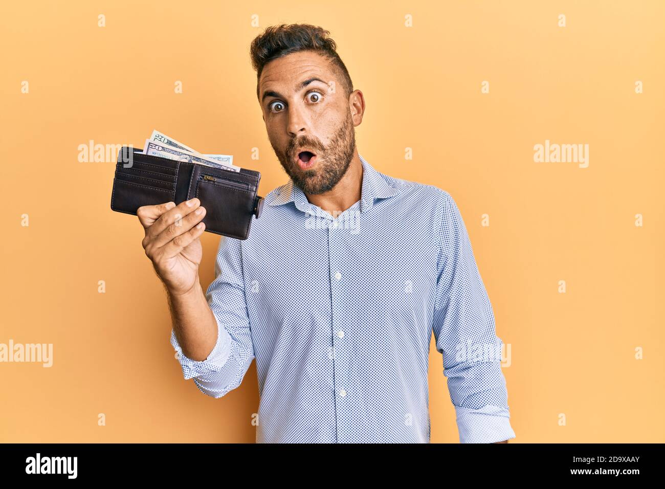 Handsome man with beard holding wallet with dollars scared and amazed ...