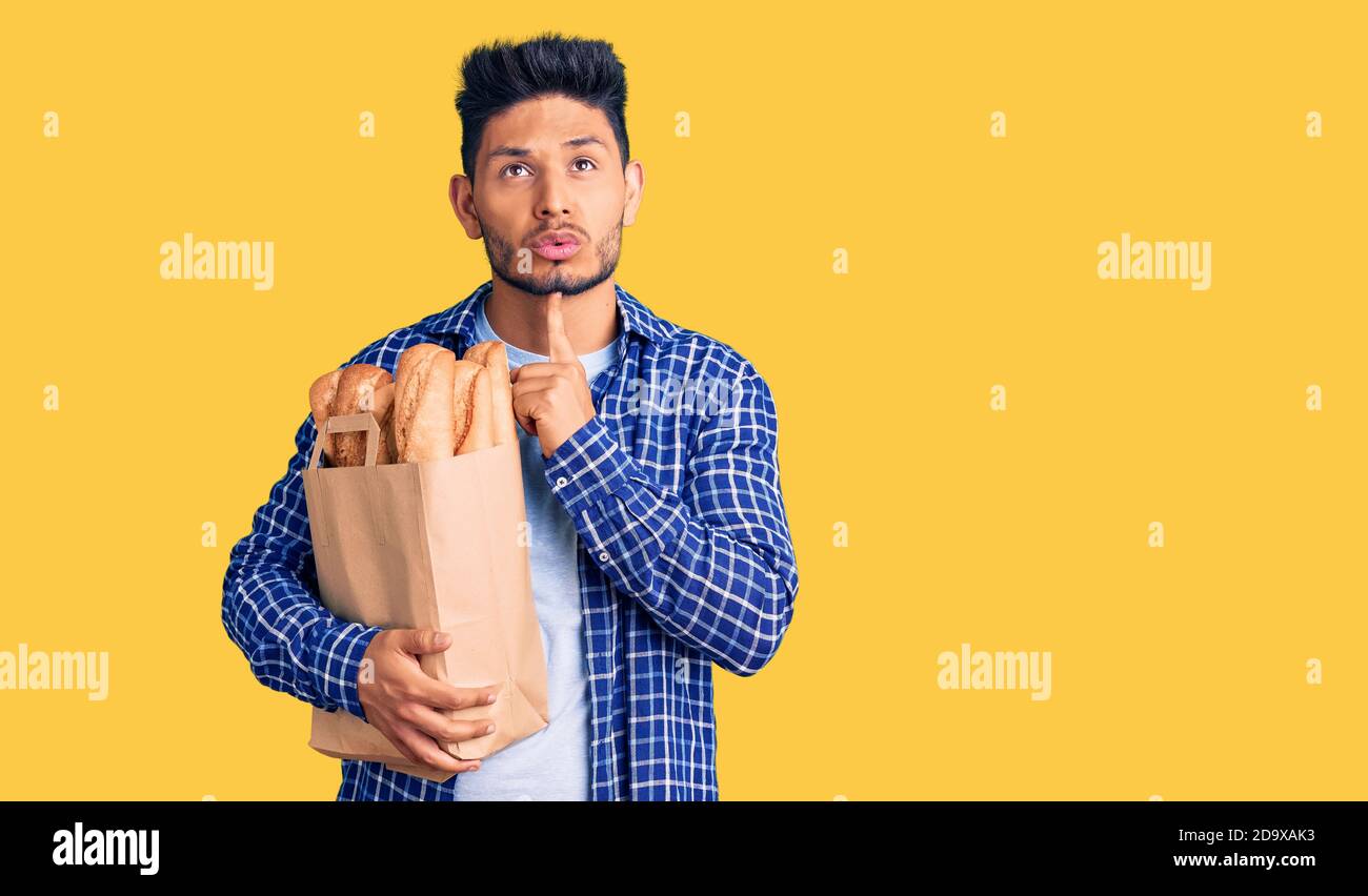 Handsome latin american young man holding paper bag with bread thinking ...