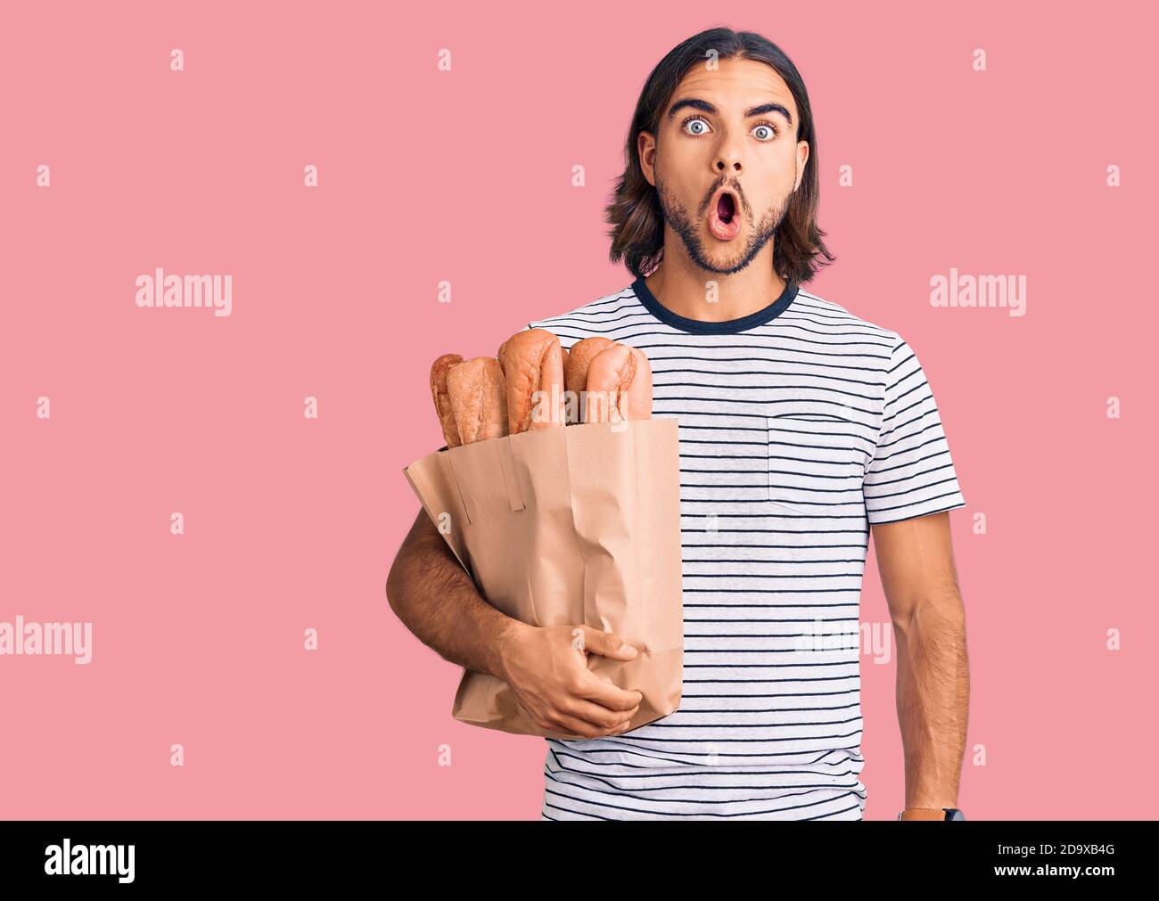Young handsome man holding paper bag with bread scared and amazed with ...