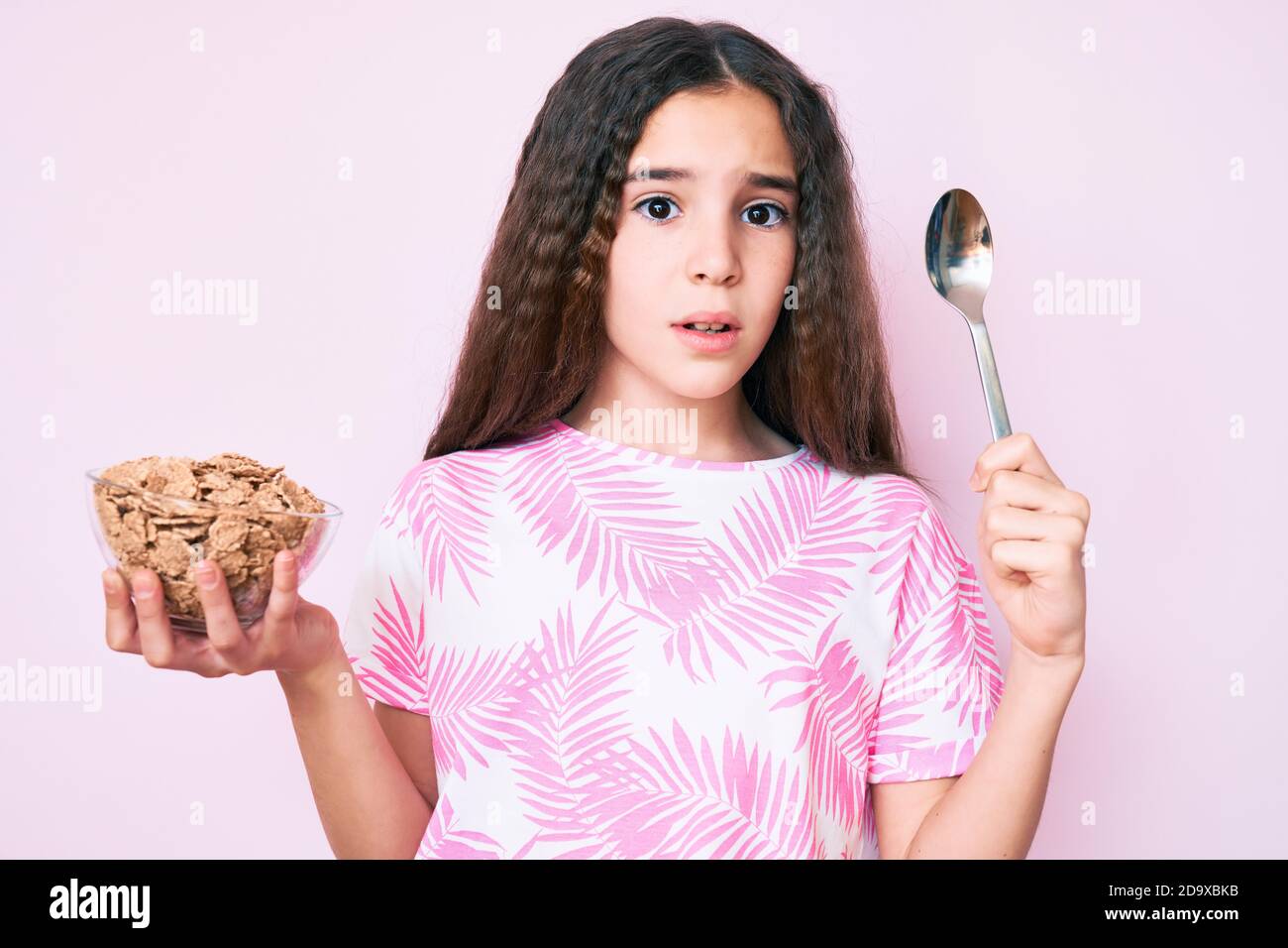Cute hispanic girl holding cornflakes and spoon clueless and confused ...