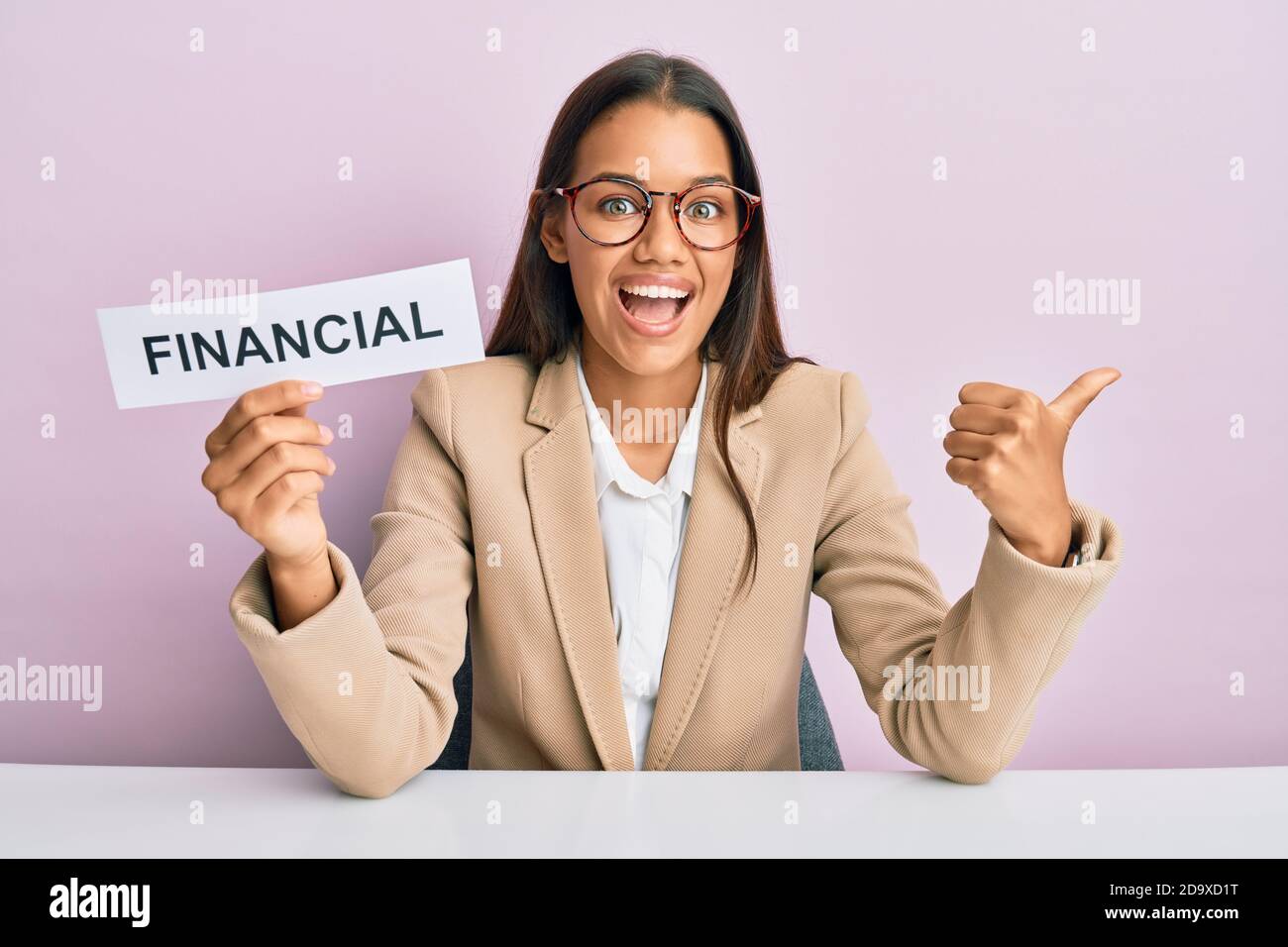 Beautiful hispanic business woman holding financial message paper pointing thumb up to the side ...