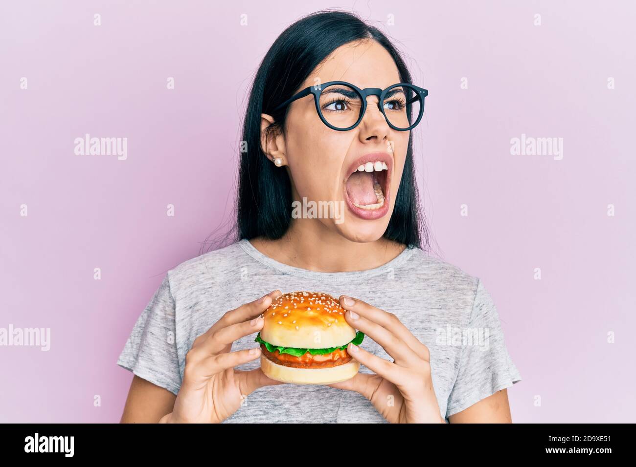 Beautiful young woman eating tasty hamburger angry and mad screaming ...