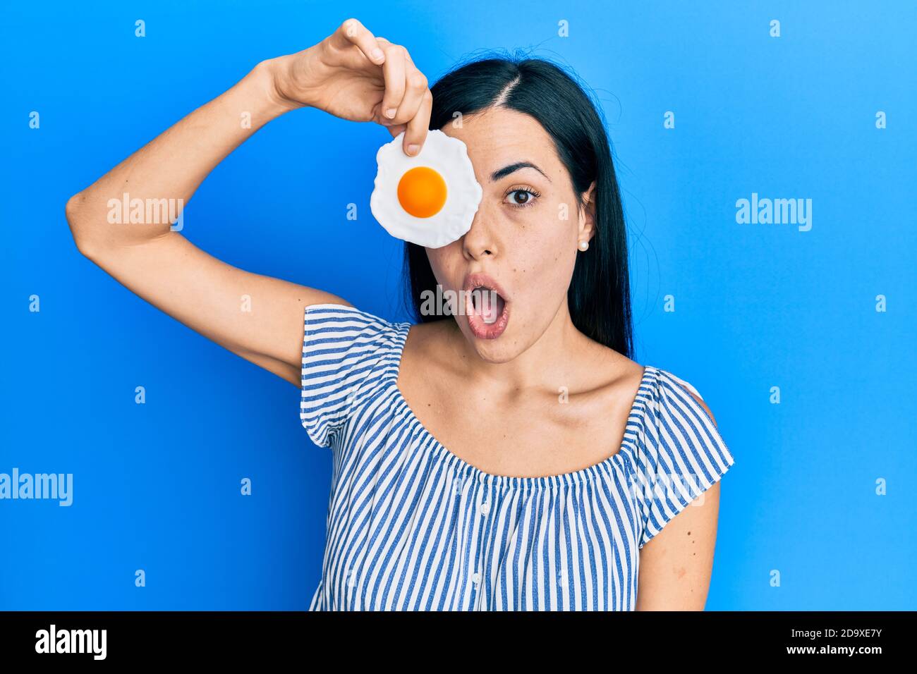 Beautiful young woman holding fried egg scared and amazed with open ...