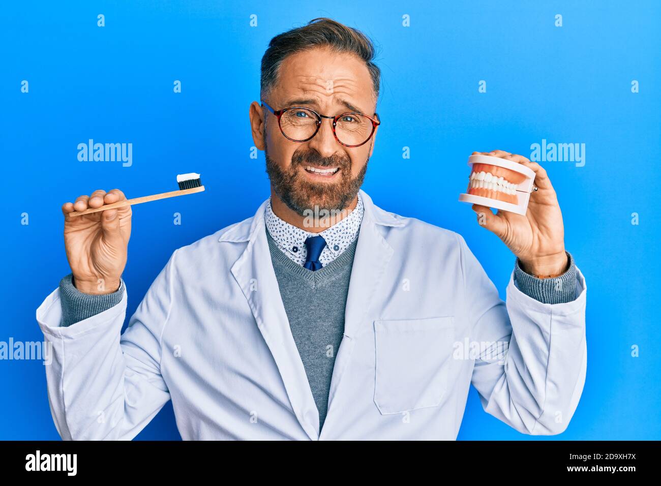 Handsome middle age dentist man holding denture and toothbrush clueless ...