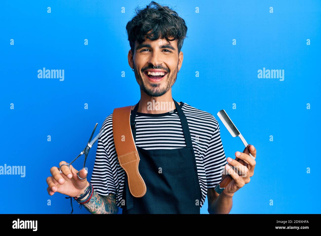 Young hispanic man wearing barber apron holding razor and scissors ...