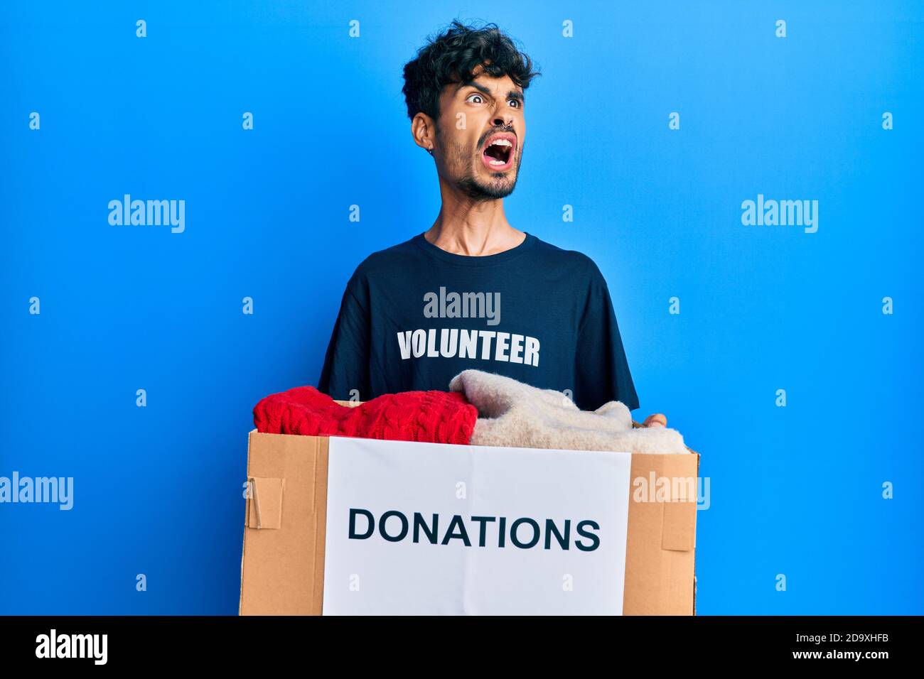 Young hispanic man holding donation box with clothes angry and mad ...