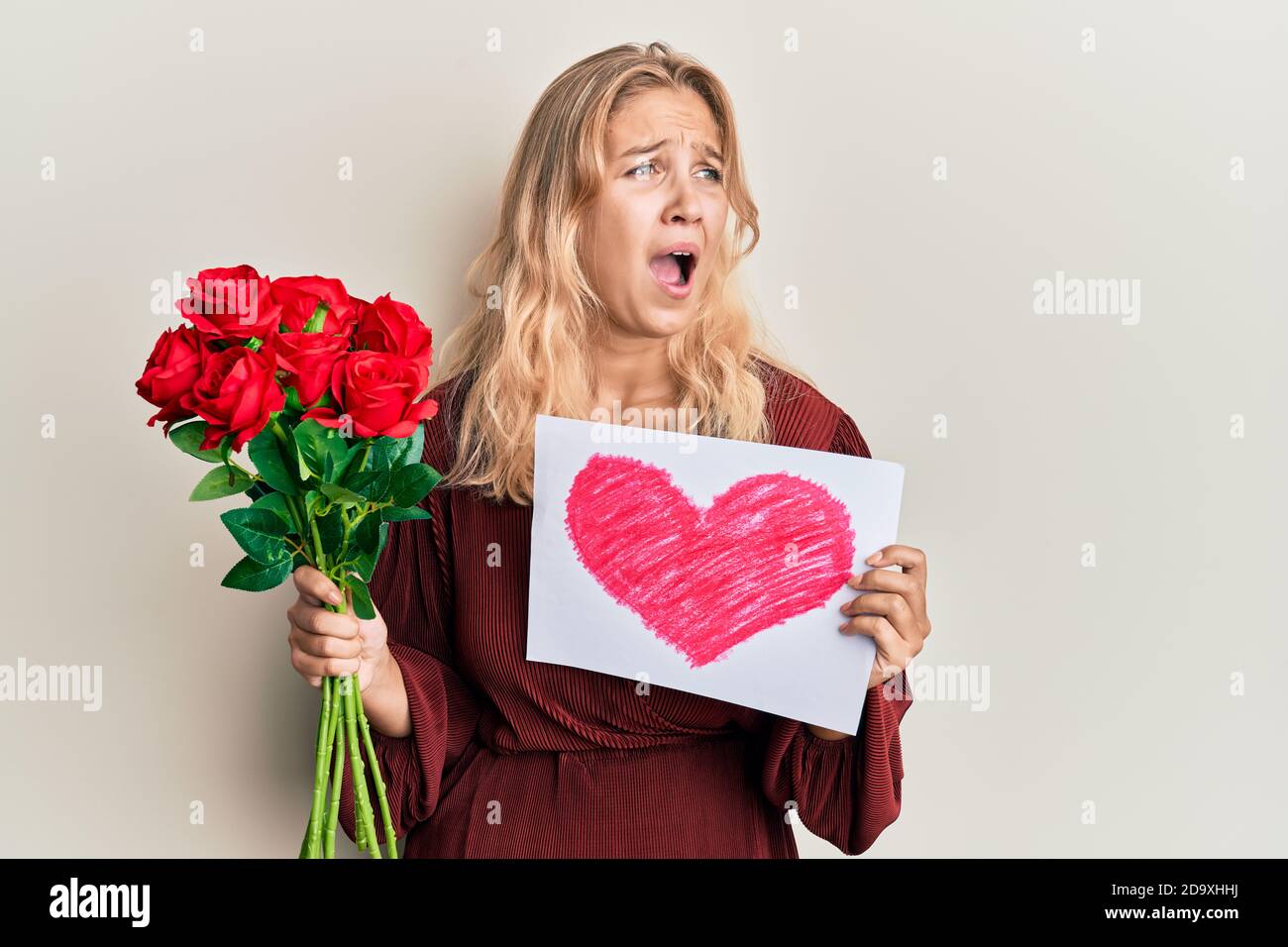 Young blonde girl holding heart draw and bouquet of roses angry and mad ...