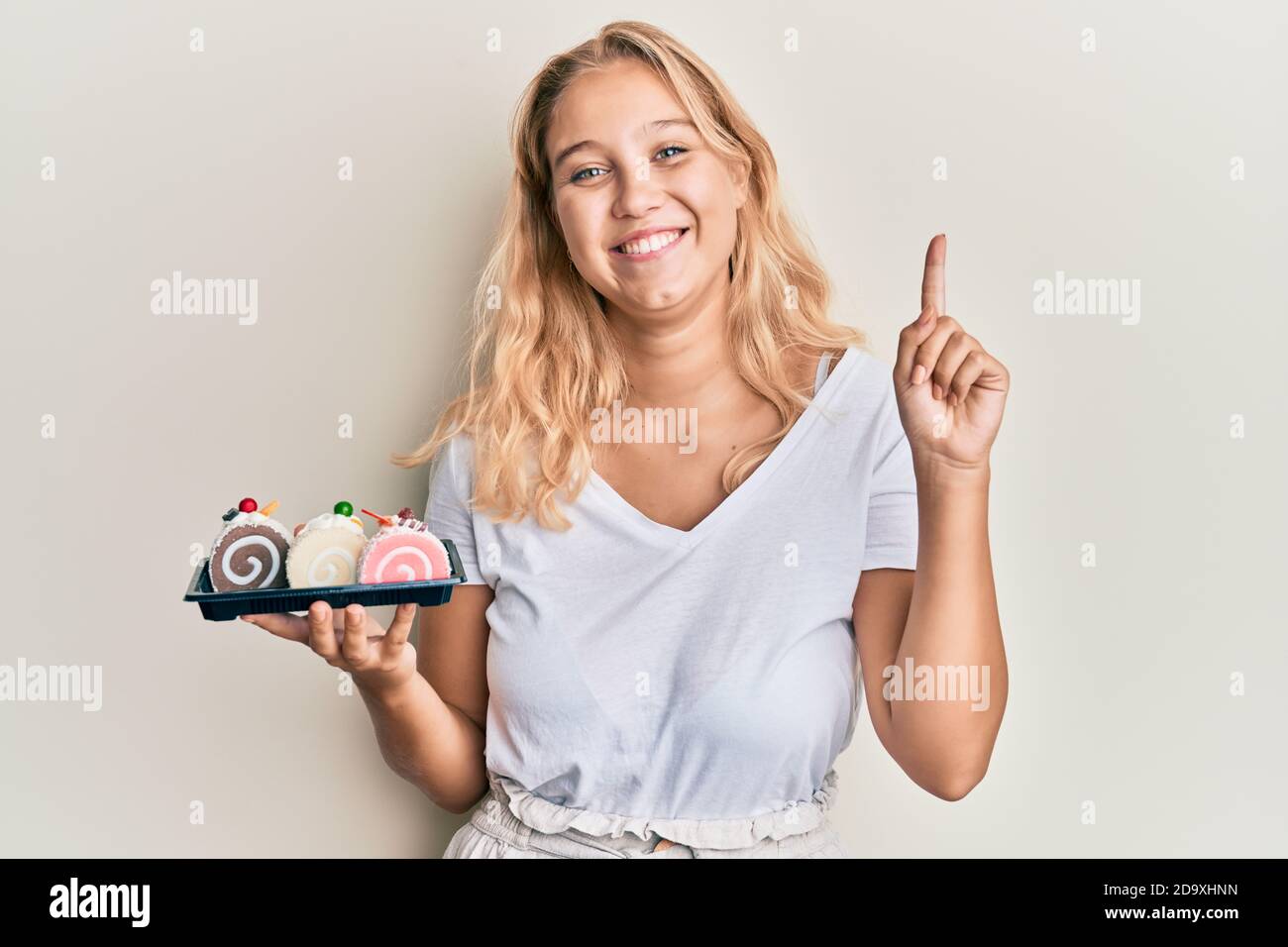 Young blonde girl holding cake sweets smiling with an idea or question ...