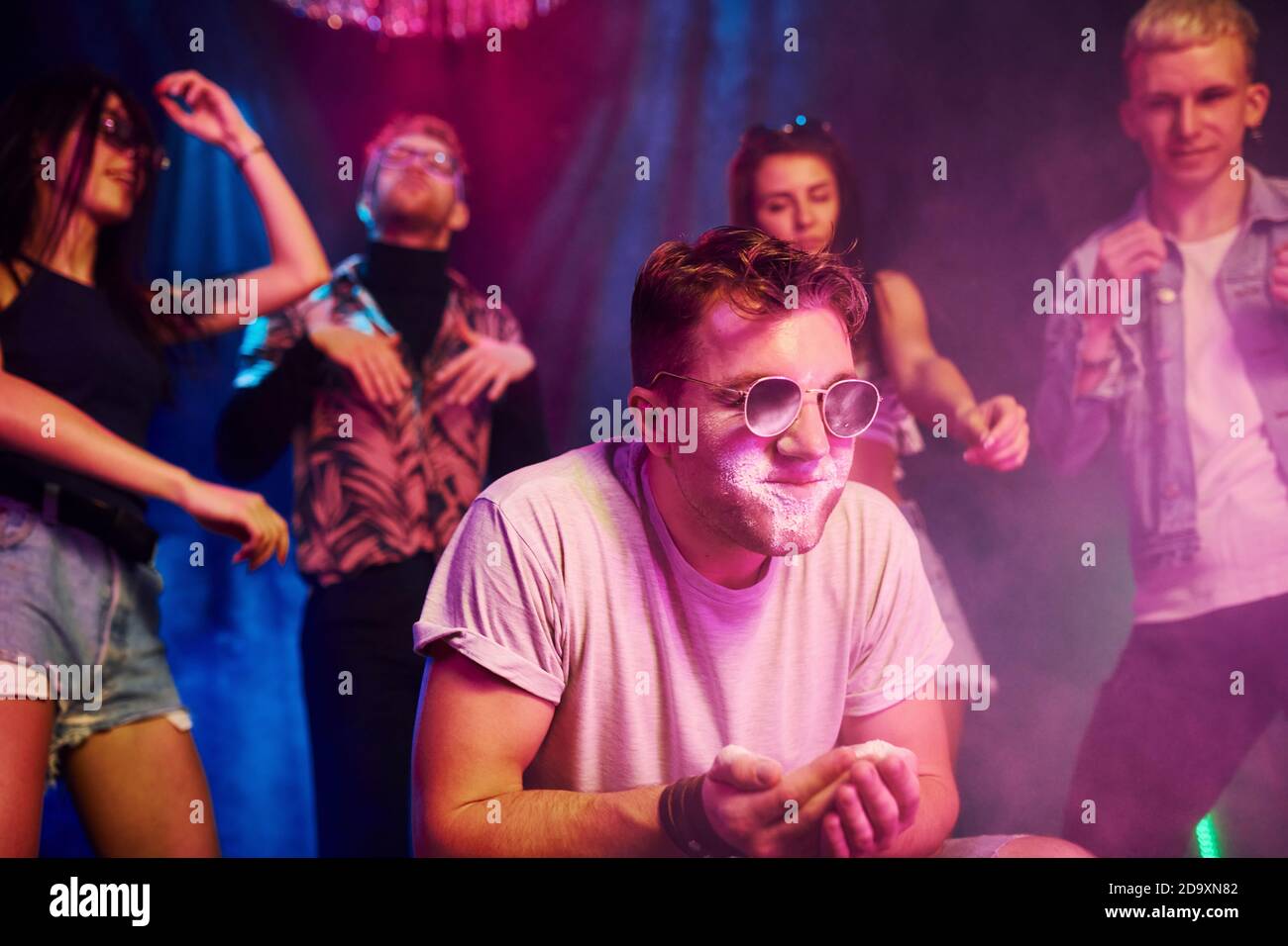 Young guy in sunglasses sniffing drug in night club with friends behind ...
