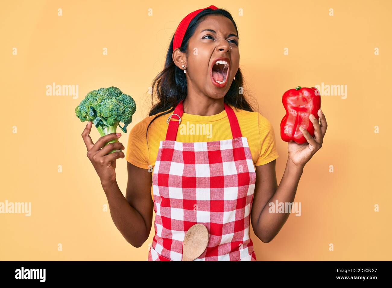 Young indian girl wearing apron holding broccoli and red pepper angry ...