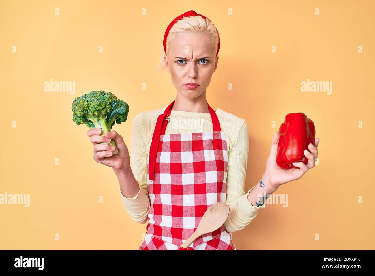 Young blonde woman with tattoo wearing cook apron holding broccoli and ...