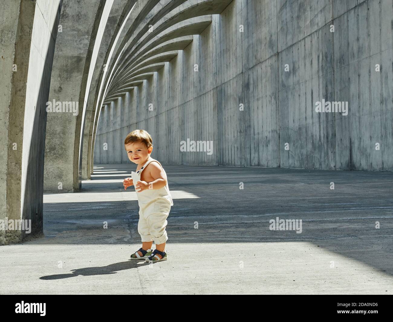 Delighted little kid in cute overalls walking along arched pathway in ...
