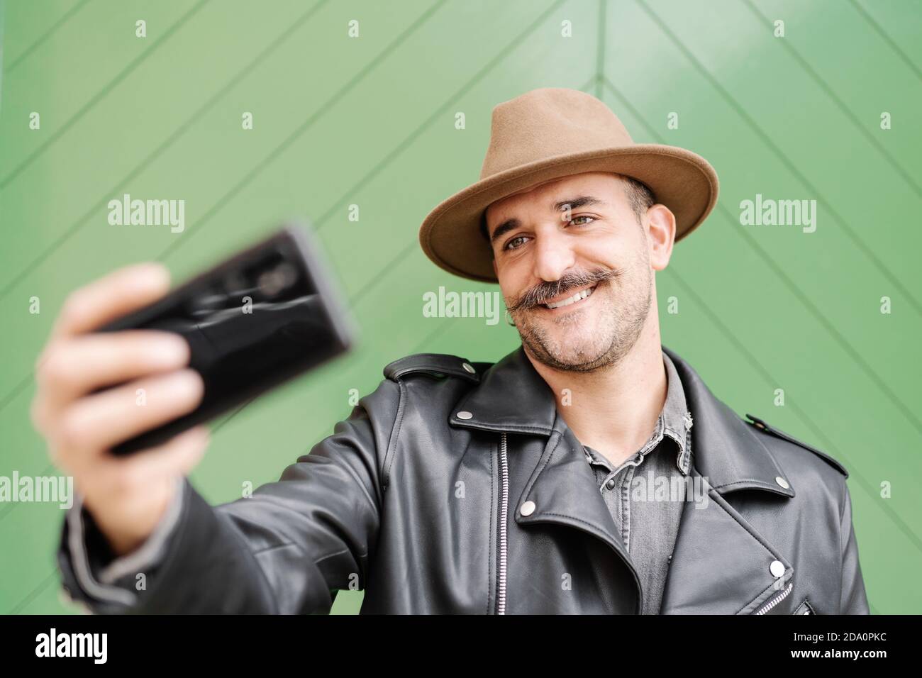 Cheerful glad bearded gallant male in hat taking photo in yard of green ...