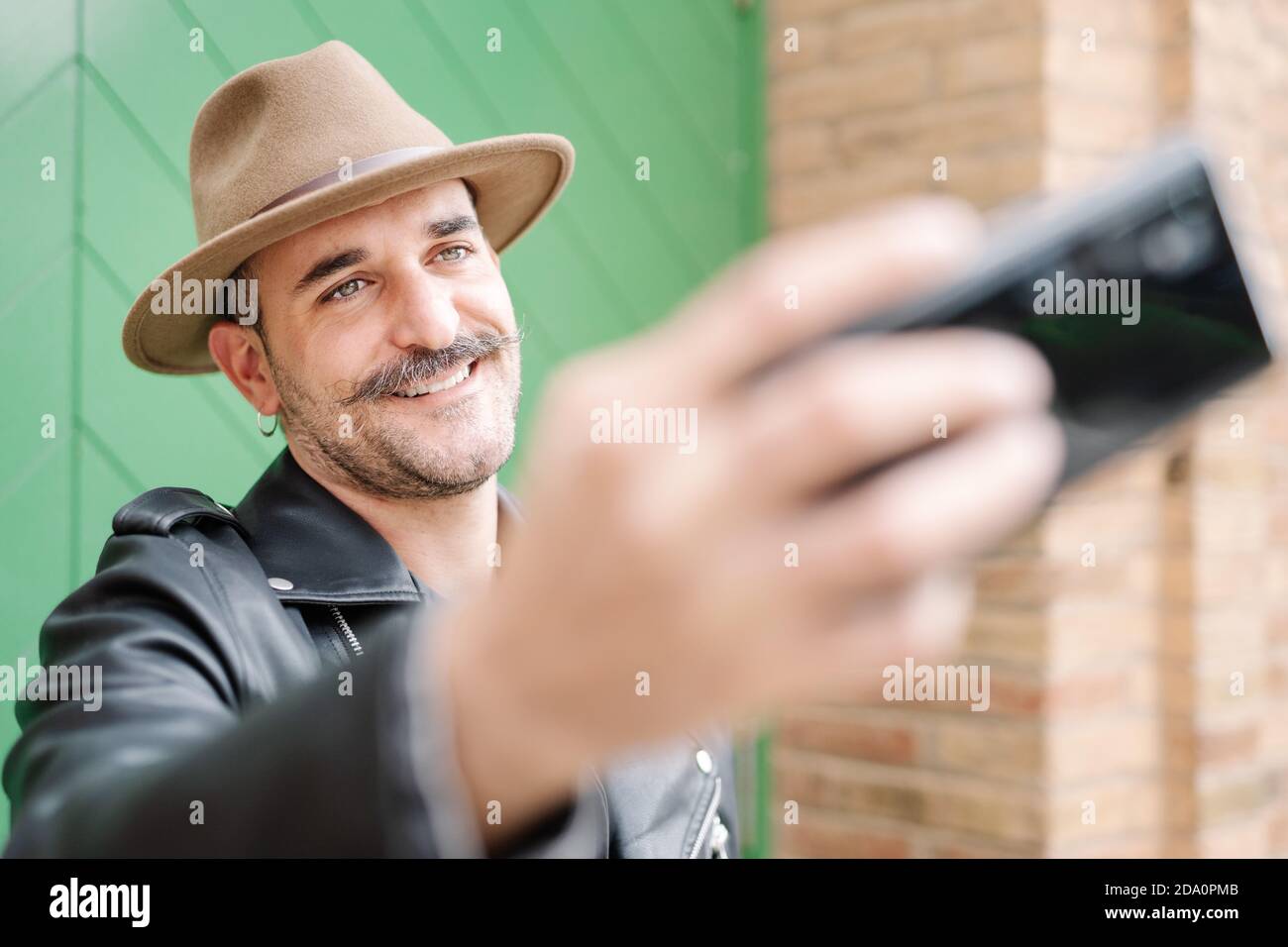 Cheerful glad bearded gallant male in hat taking photo in yard of brick ...