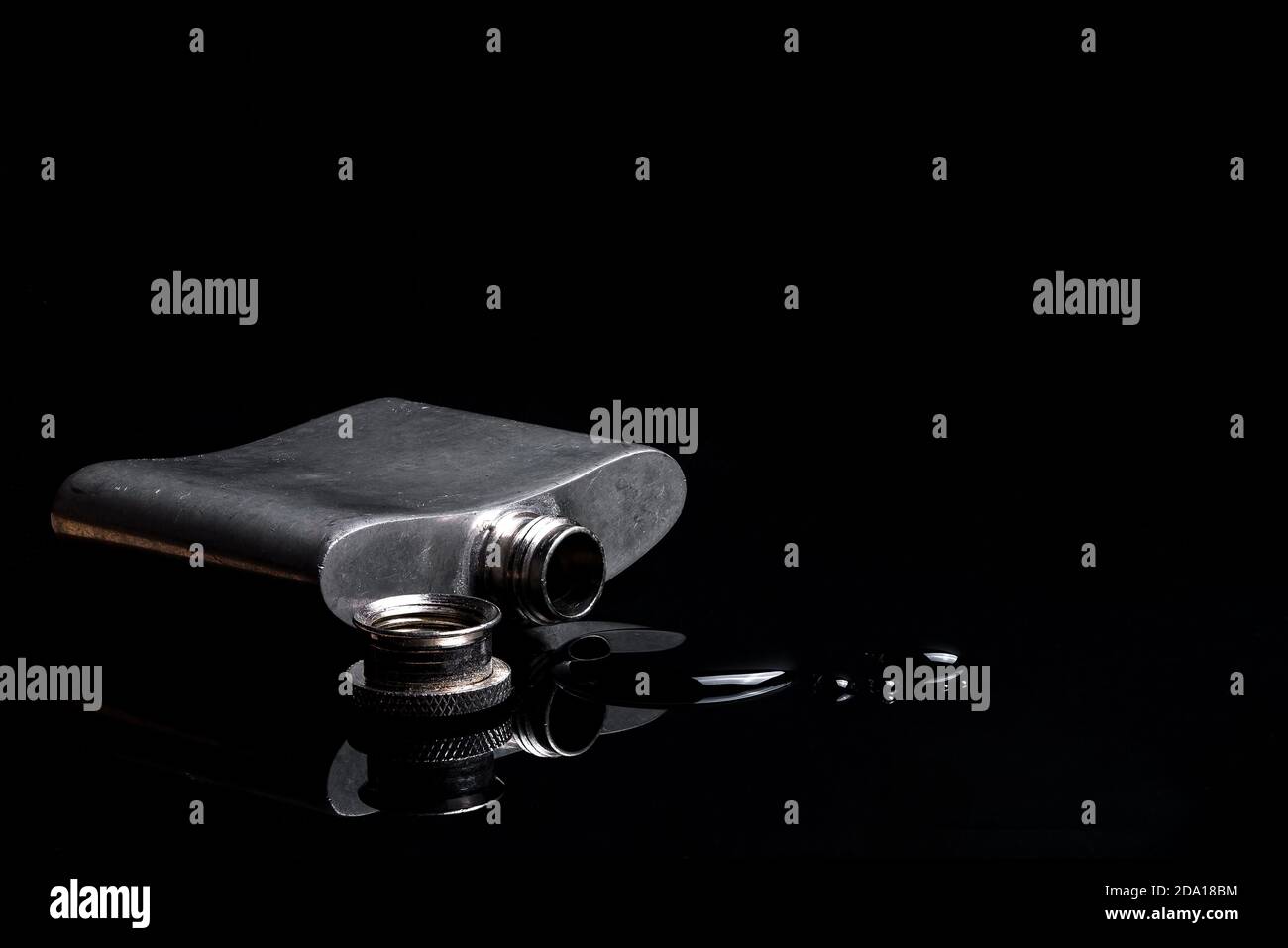 Small worn metallic hip flask spilled over on a dark reflective surface ...