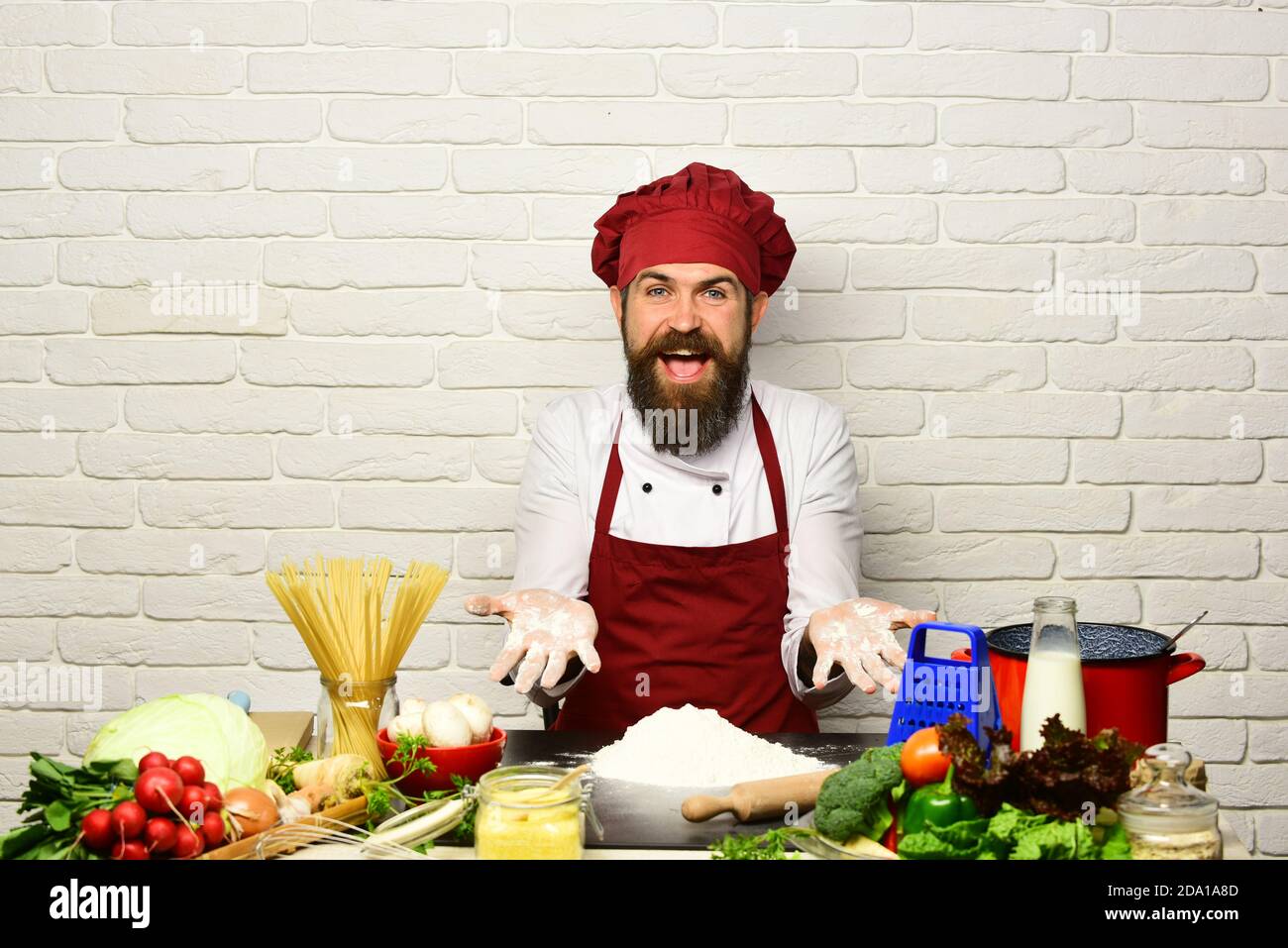 Cook with happy face in uniform sits by kitchen table with vegetables ...