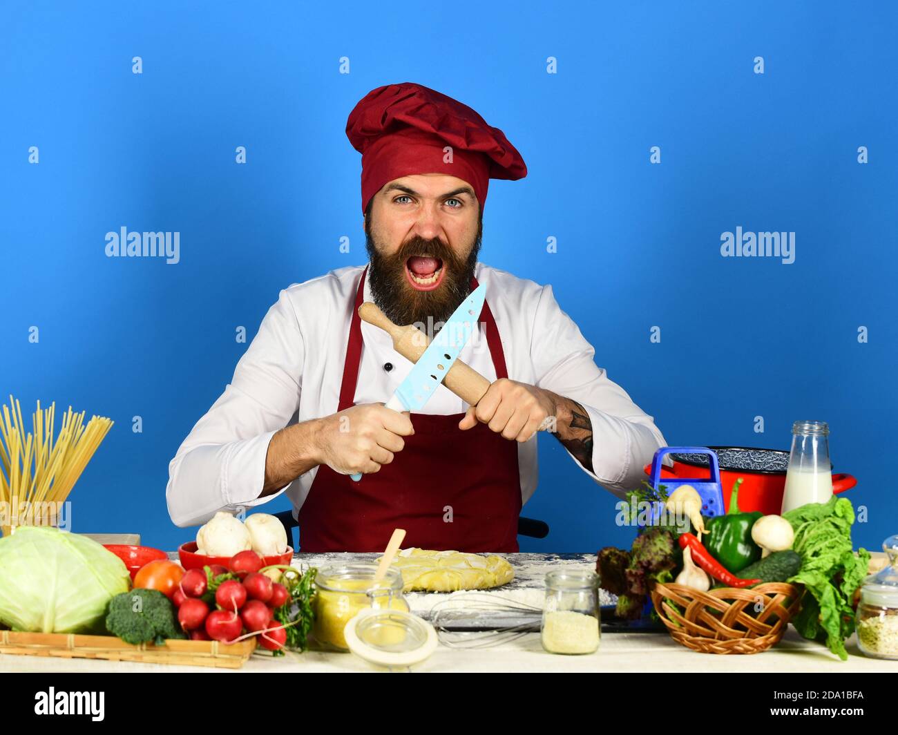 Chef with pasta, vegetables and dough on table. Cook with angry face in ...