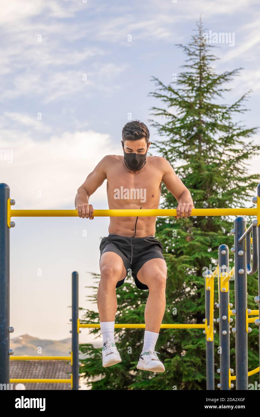 Latin young man without a shirt and black pants and mask doing pull-ups ...