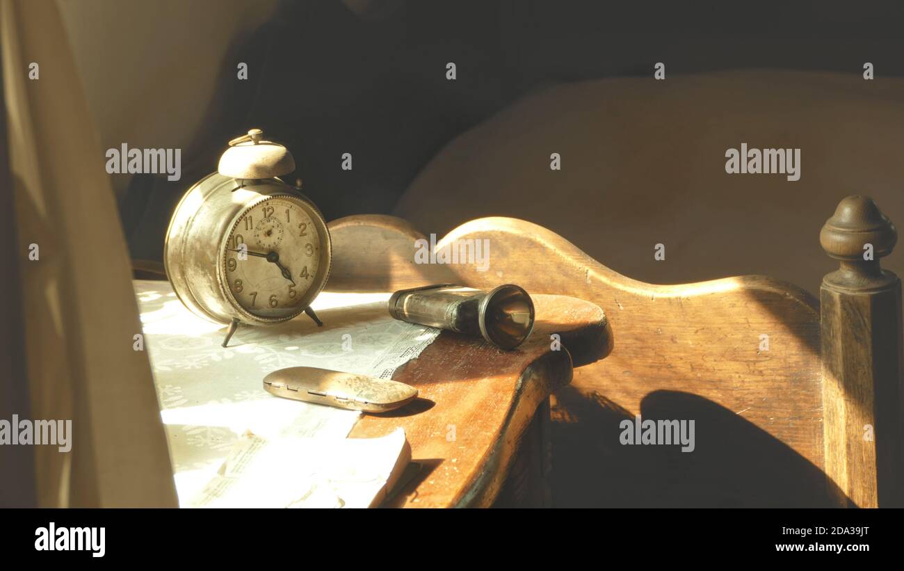 Antique alarm clock on wooden bedside table with bed in the background ...