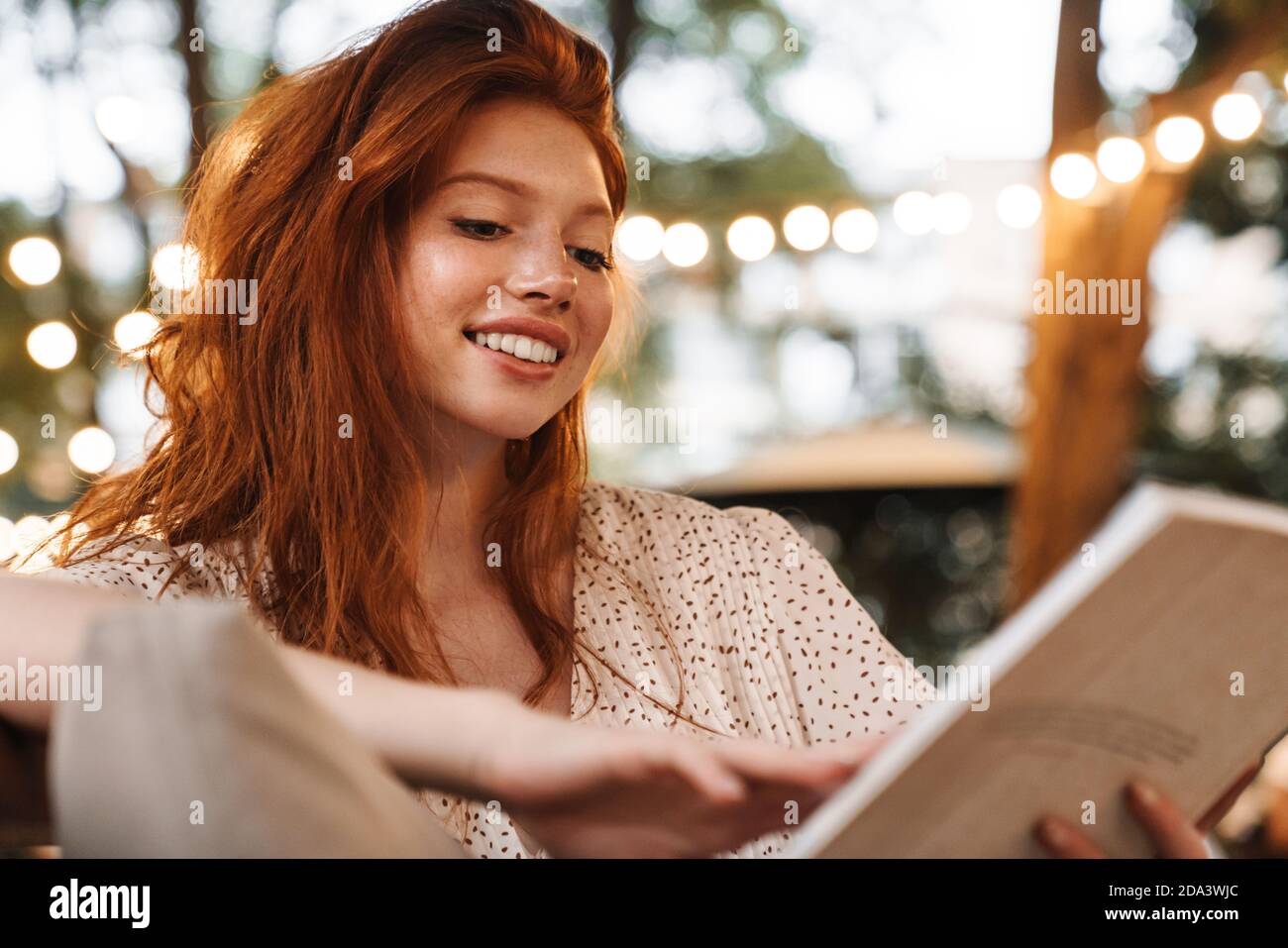 Image of joyful ginger girl smiling and reading book while sitting in ...