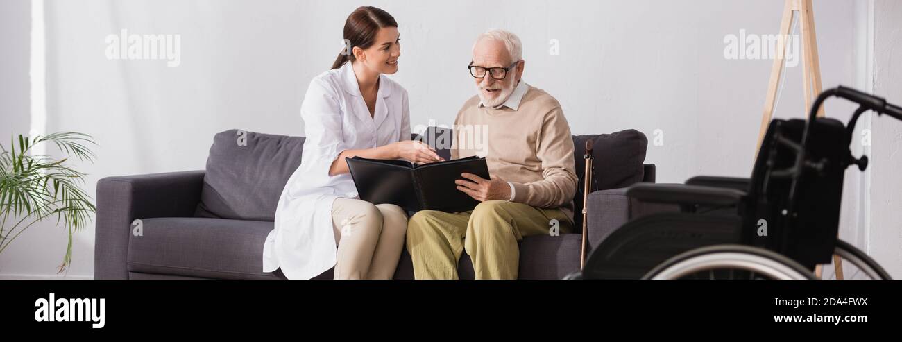 geriatric nurse and elderly man looking at photos while sitting on sofa ...