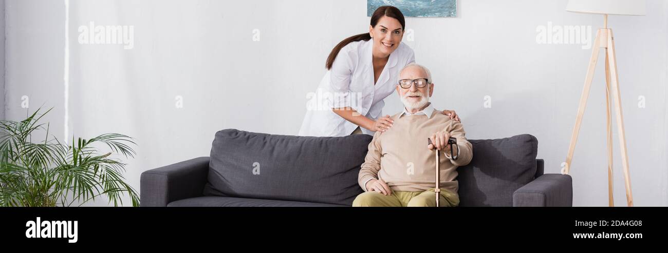 geriatric nurse and aged man smiling at camera together at home, banner ...