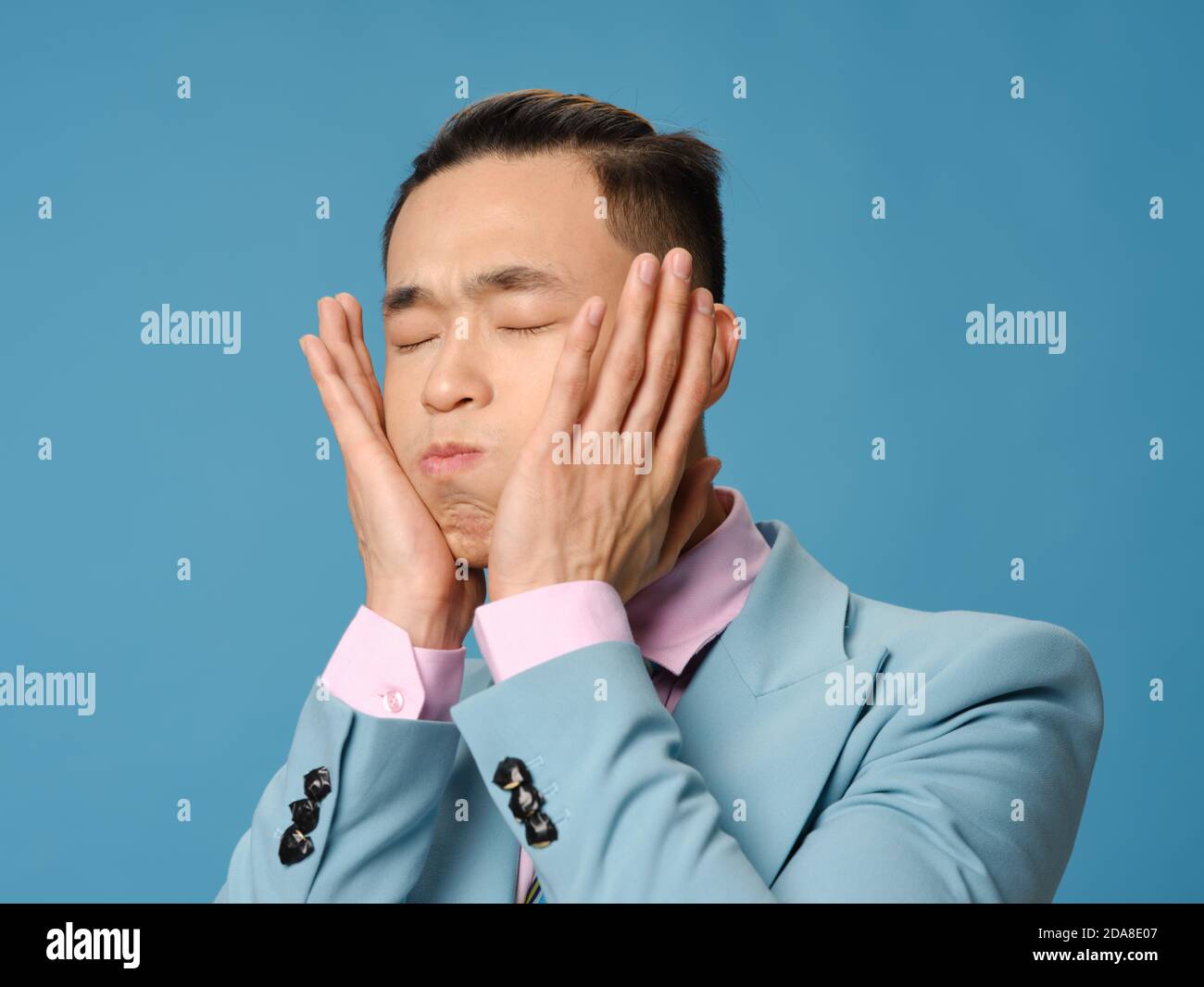 Asian man with puffy cheeks touching face with hands on blue background ...