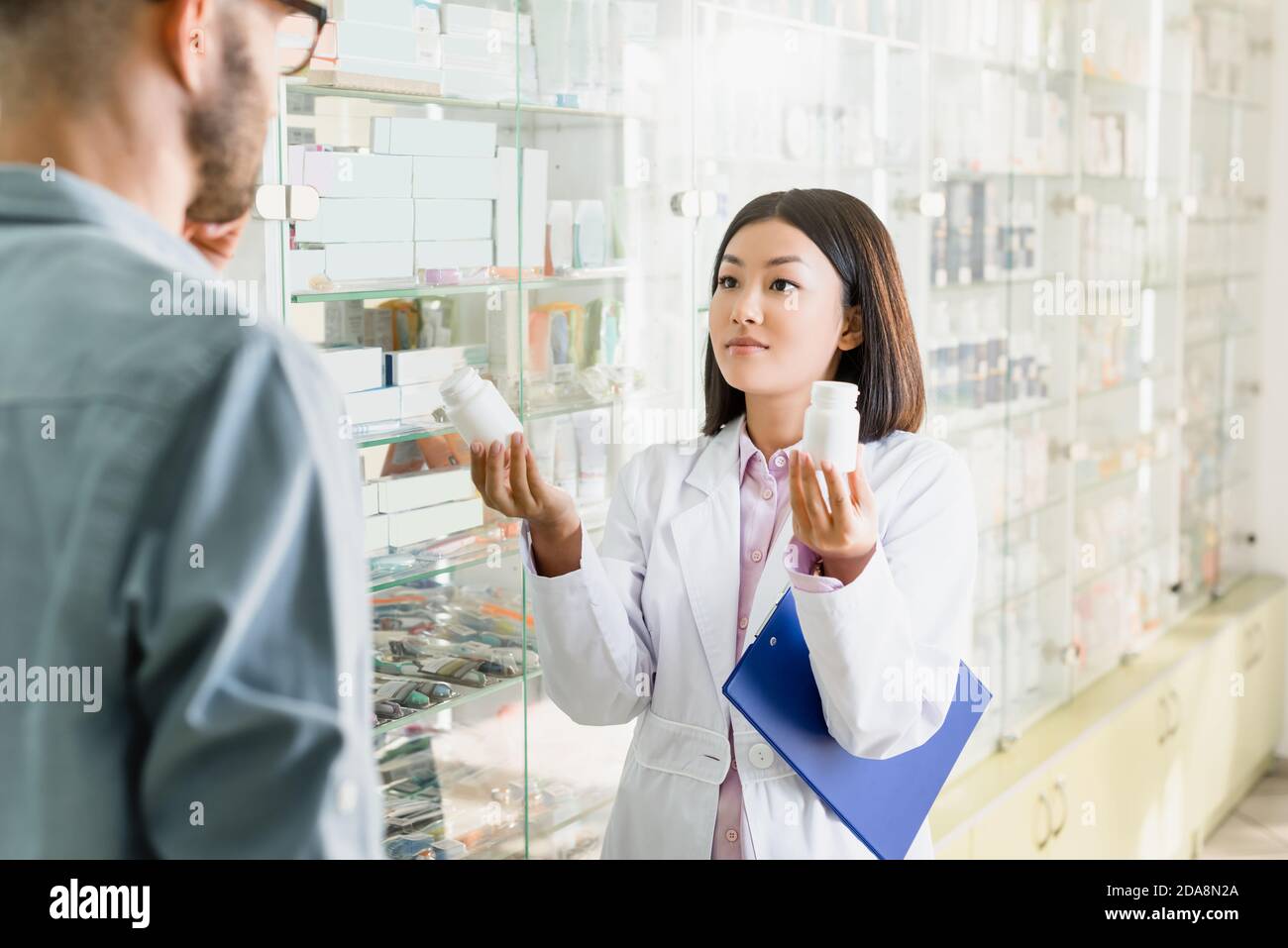 asian pharmacist in white coat holding clipboard and bottles with ...