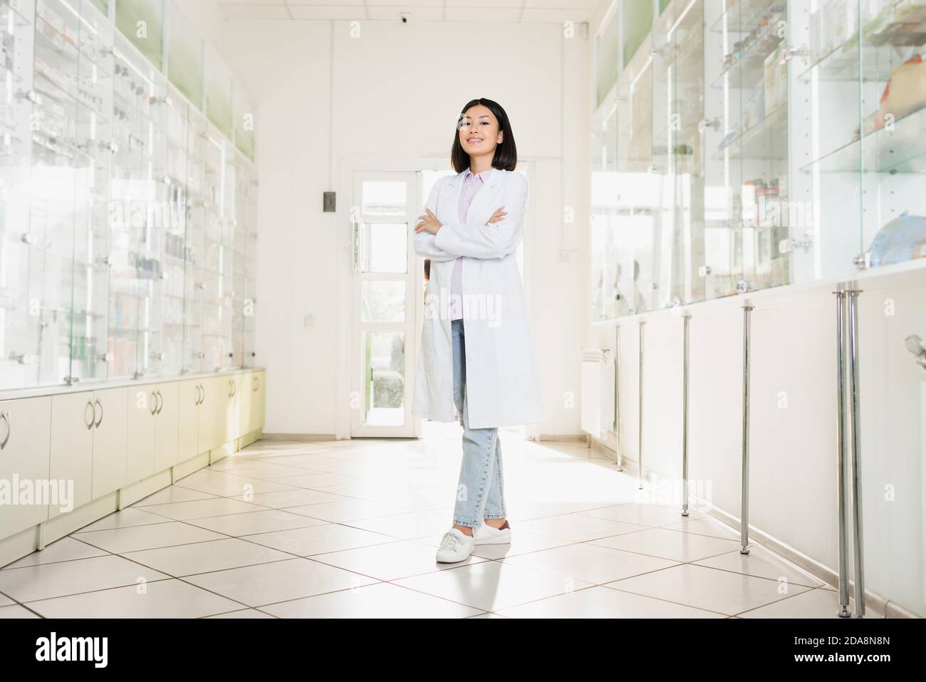 full length of smiling asian pharmacist in white coat standing with ...