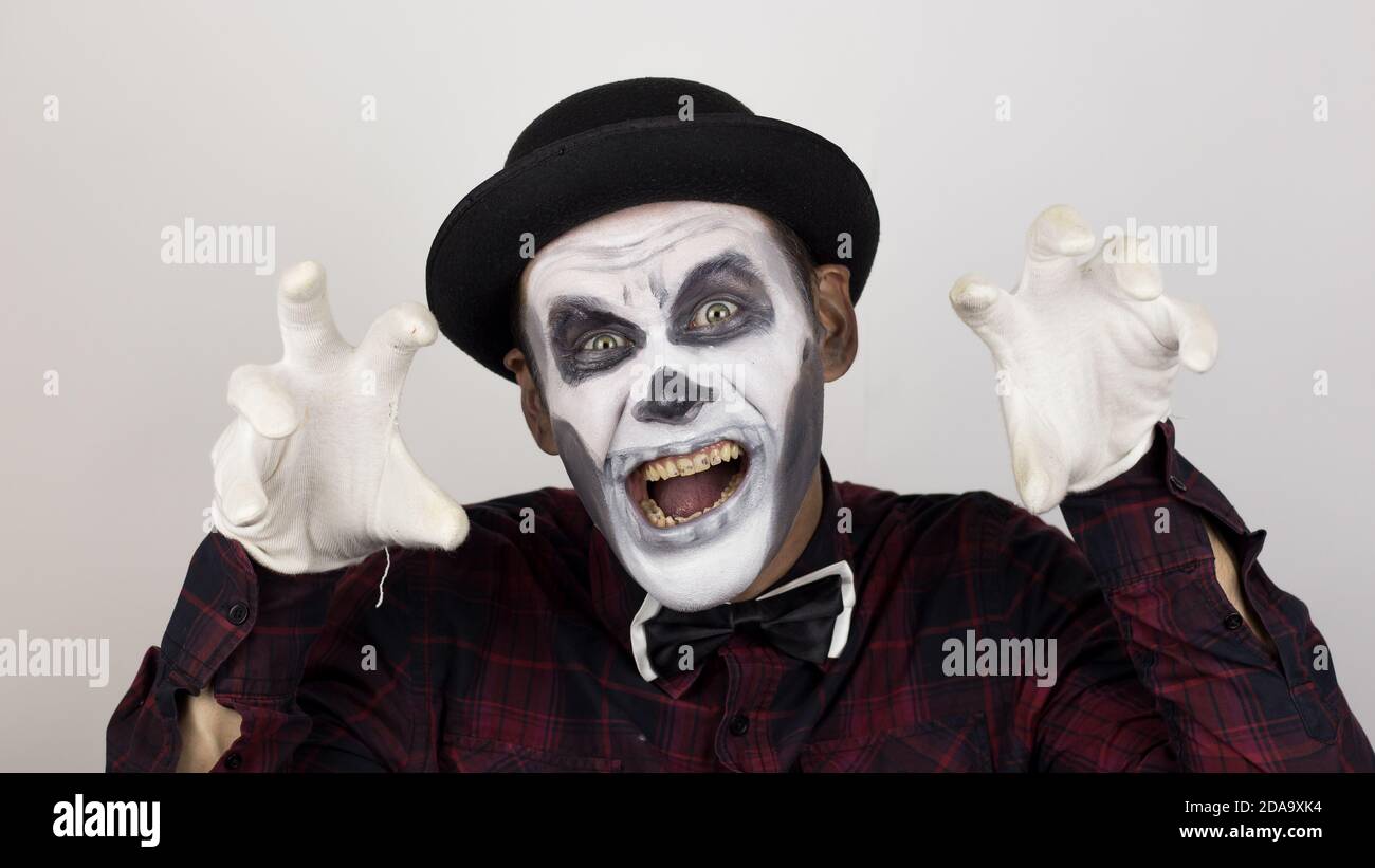 A horrible man in clown makeup grimaces and makes frightening gestures ...