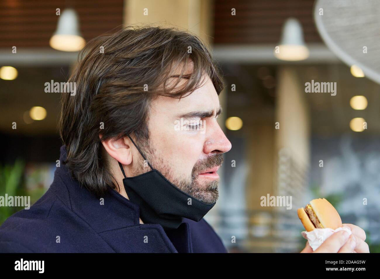 Man with a mask on his chin because of Covid-19 while eating a burger ...