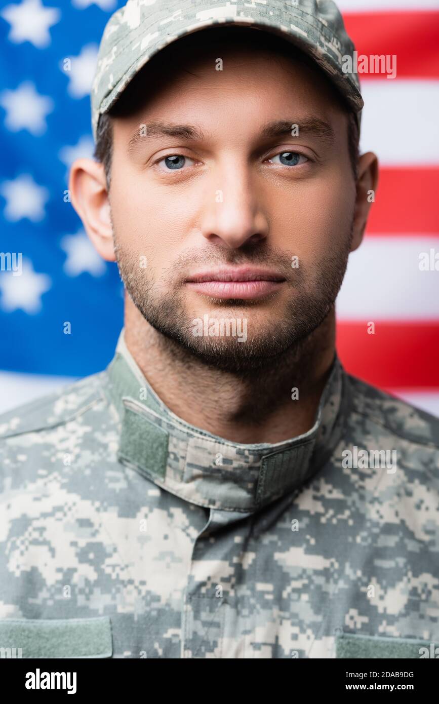 military man in uniform and cap looking at camera near american flag on ...