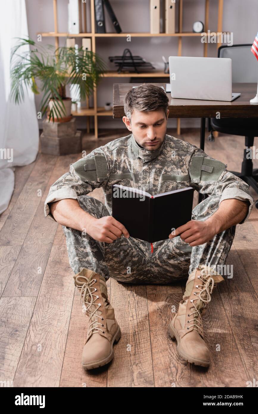 focused military man holding copy book while sitting on floor near desk ...