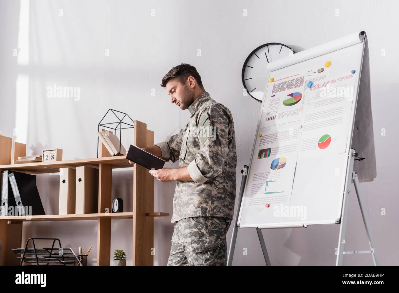 serious military man holding notepad near wooden rack and flipchart ...