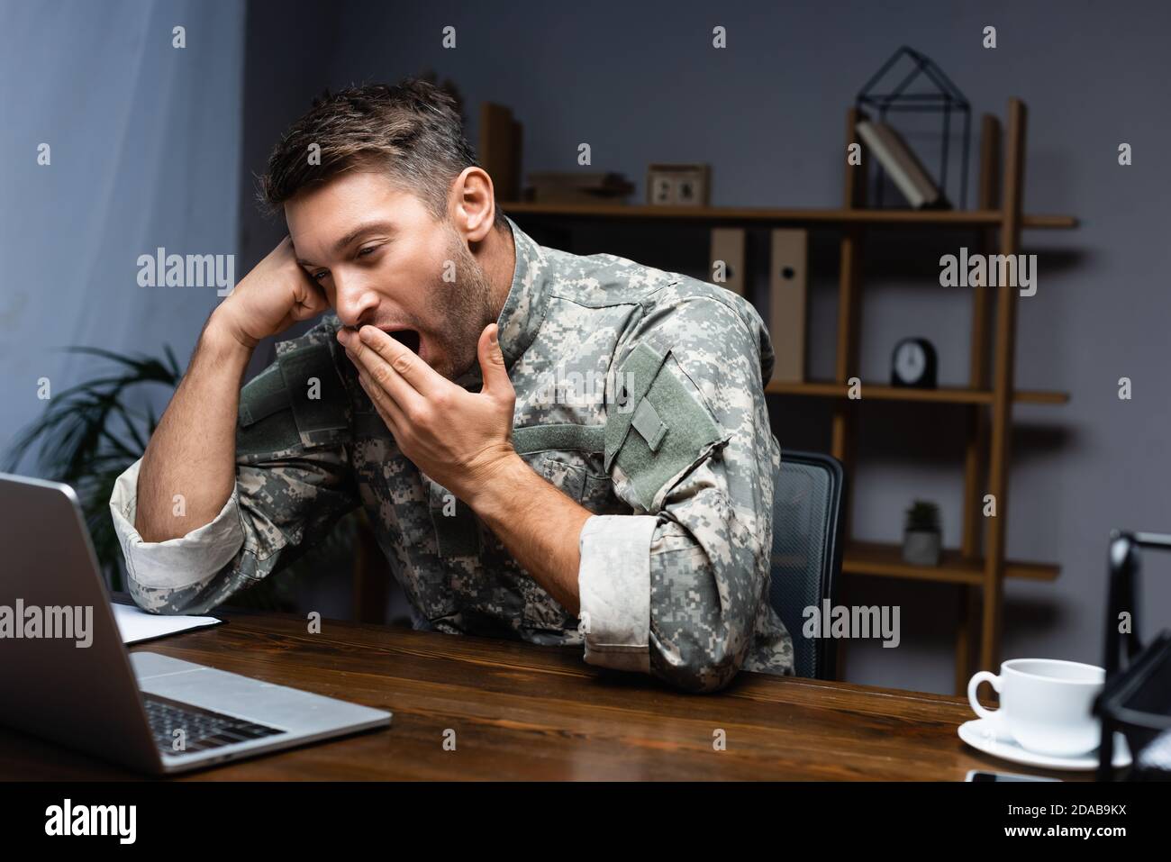 tired military man in uniform covering mouth while yawning near laptop ...