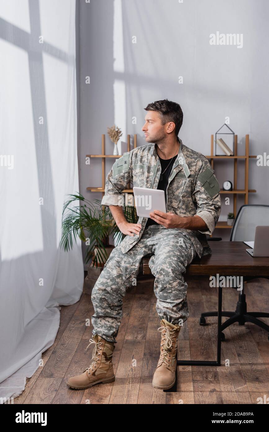 soldier in military uniform holding digital tablet near laptop on desk ...