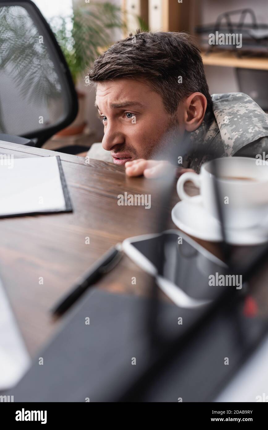 displeased military man hiding under desk with cup of coffee and ...