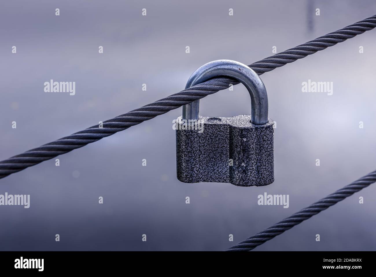 Metal padlock hanging on steel cable of bridge fence. Unfocused river ...
