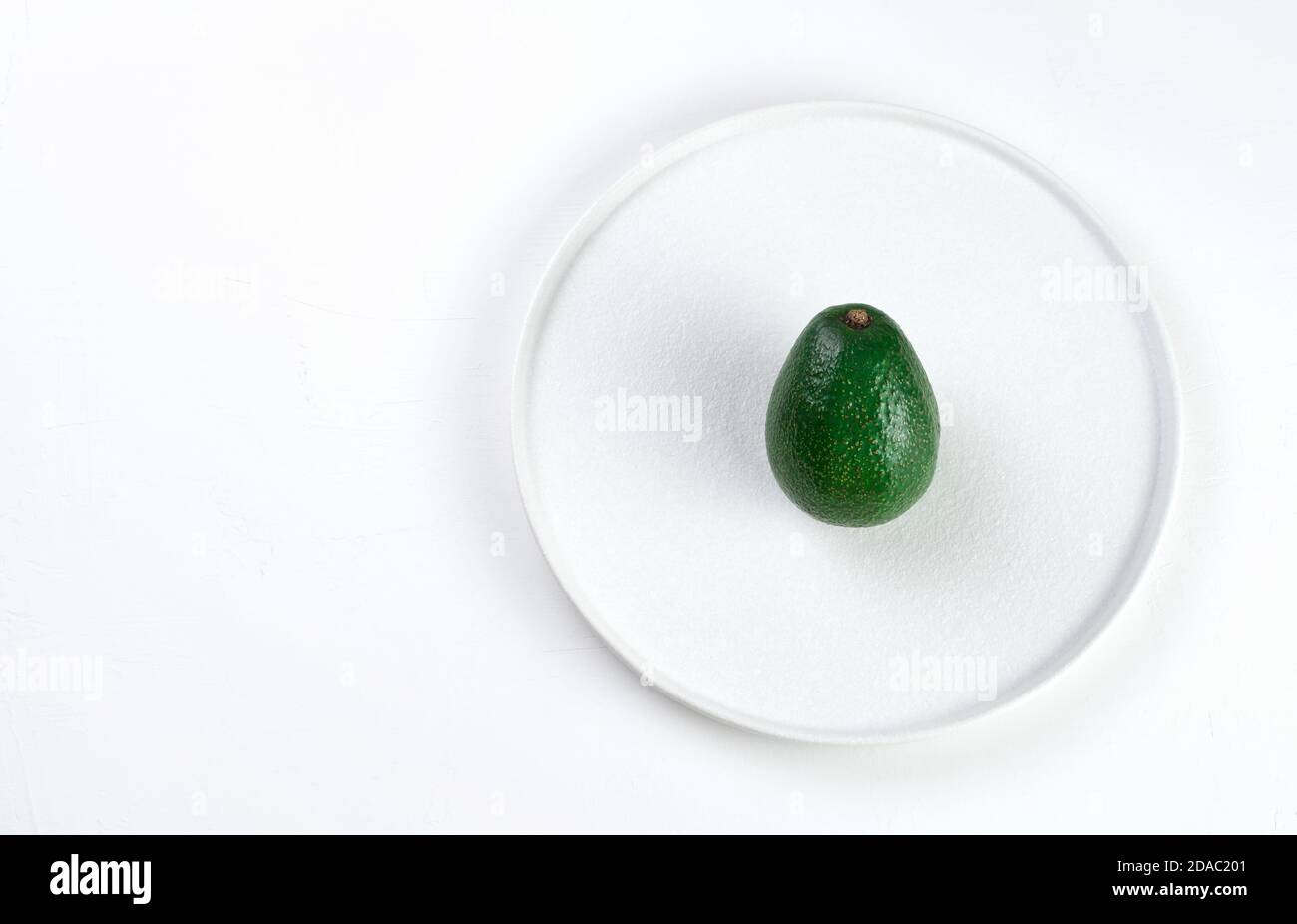 Green unpeeled avocado in a flat white plate on a light background ...