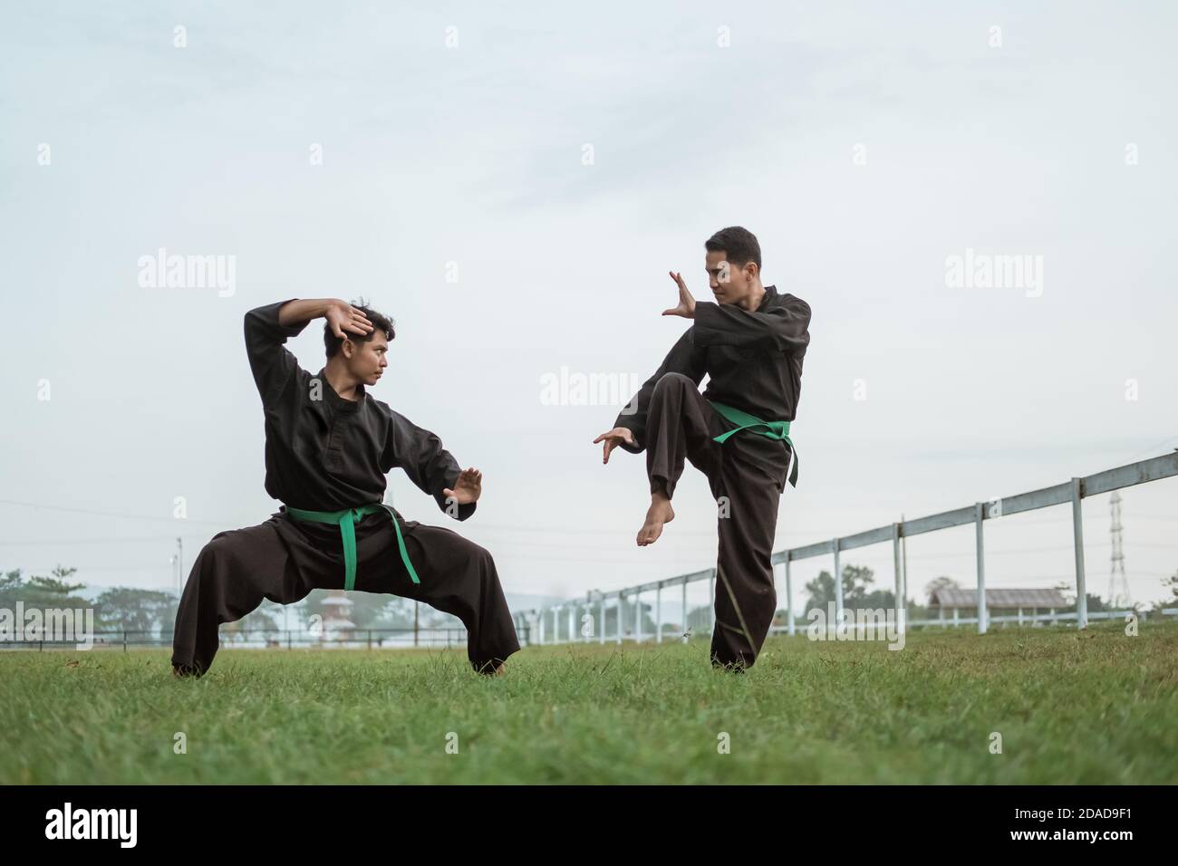 Two Asian men in pencak silat uniforms pose with a stance in a park ...