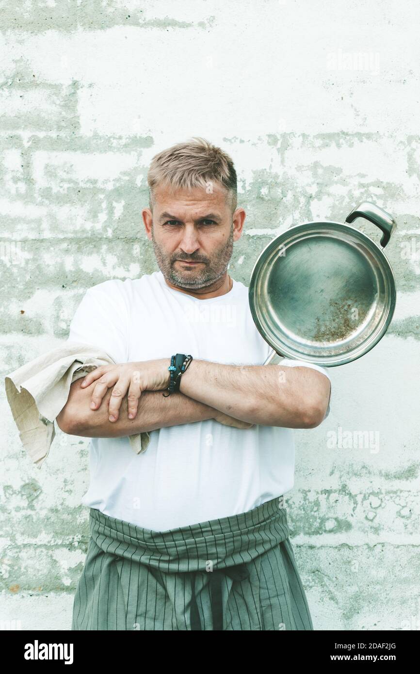 Stylish chef in an apron and with a steel frying pan on a brick wall ...