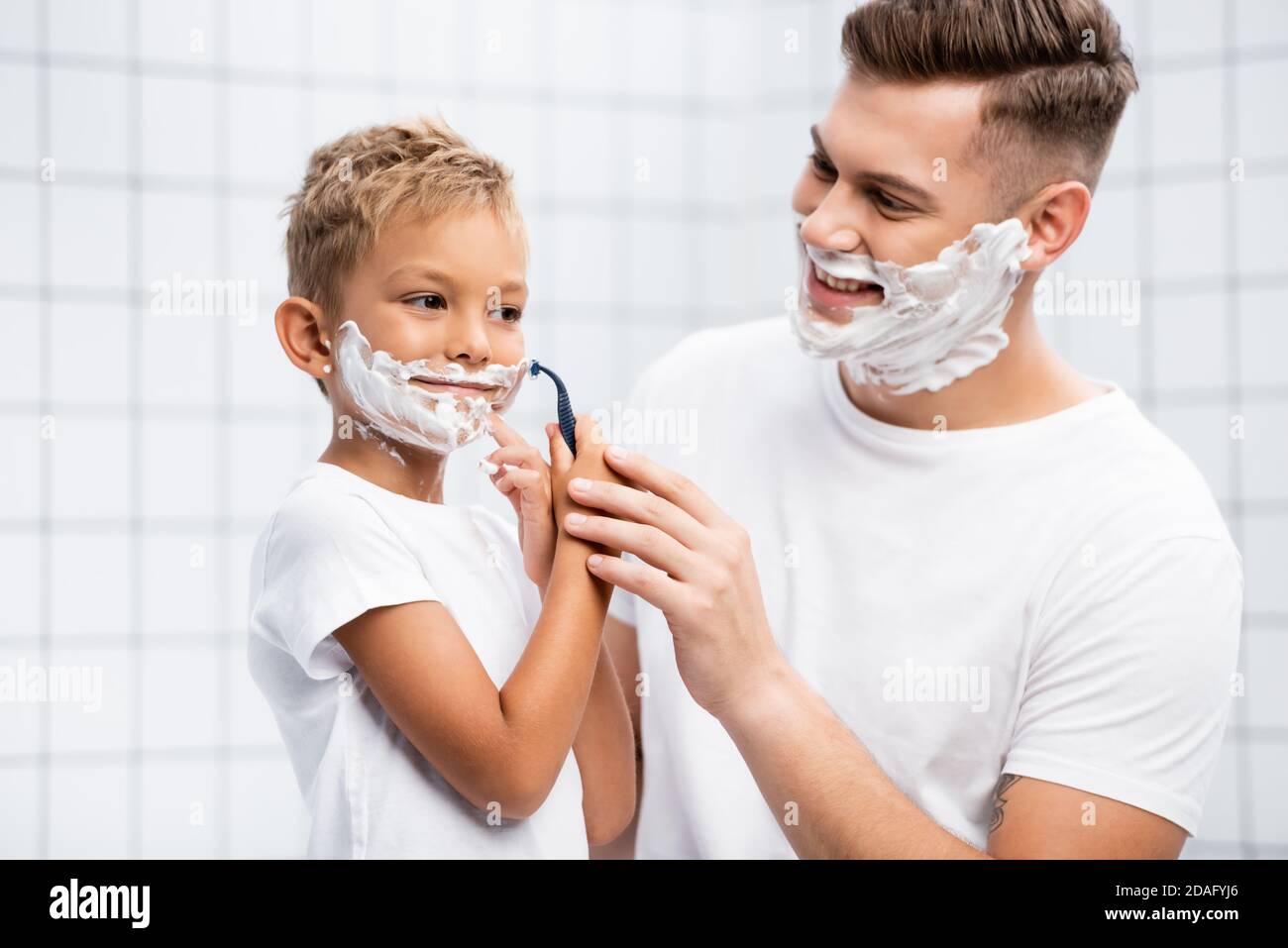 Smiling father looking at son with shaving foam on face holding safety ...