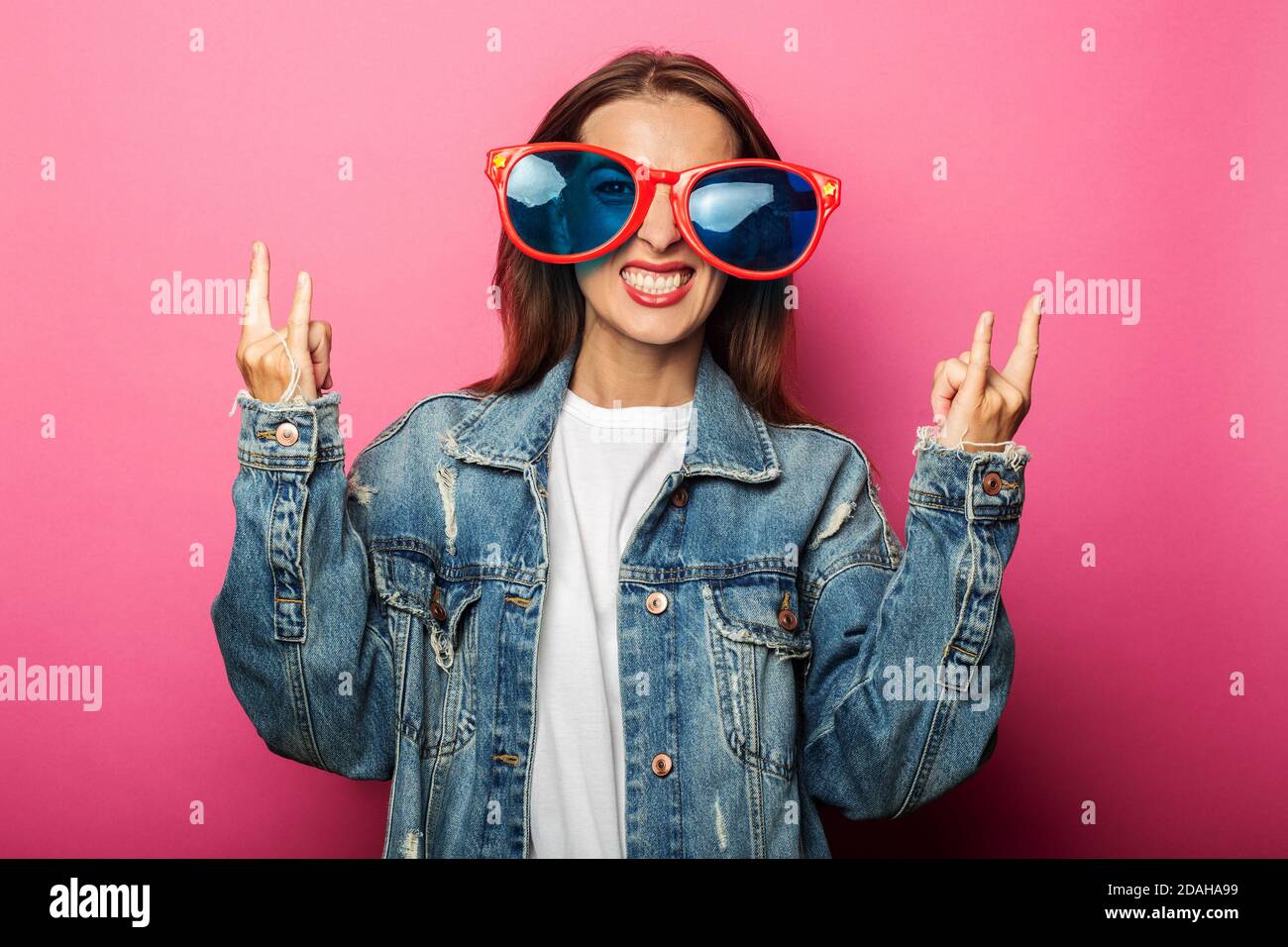 Young woman in huge glasses showing rocker horn gesture on pink ...