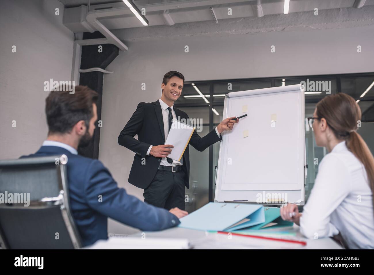 Man standing near stand and two seated colleagues Stock Photo - Alamy