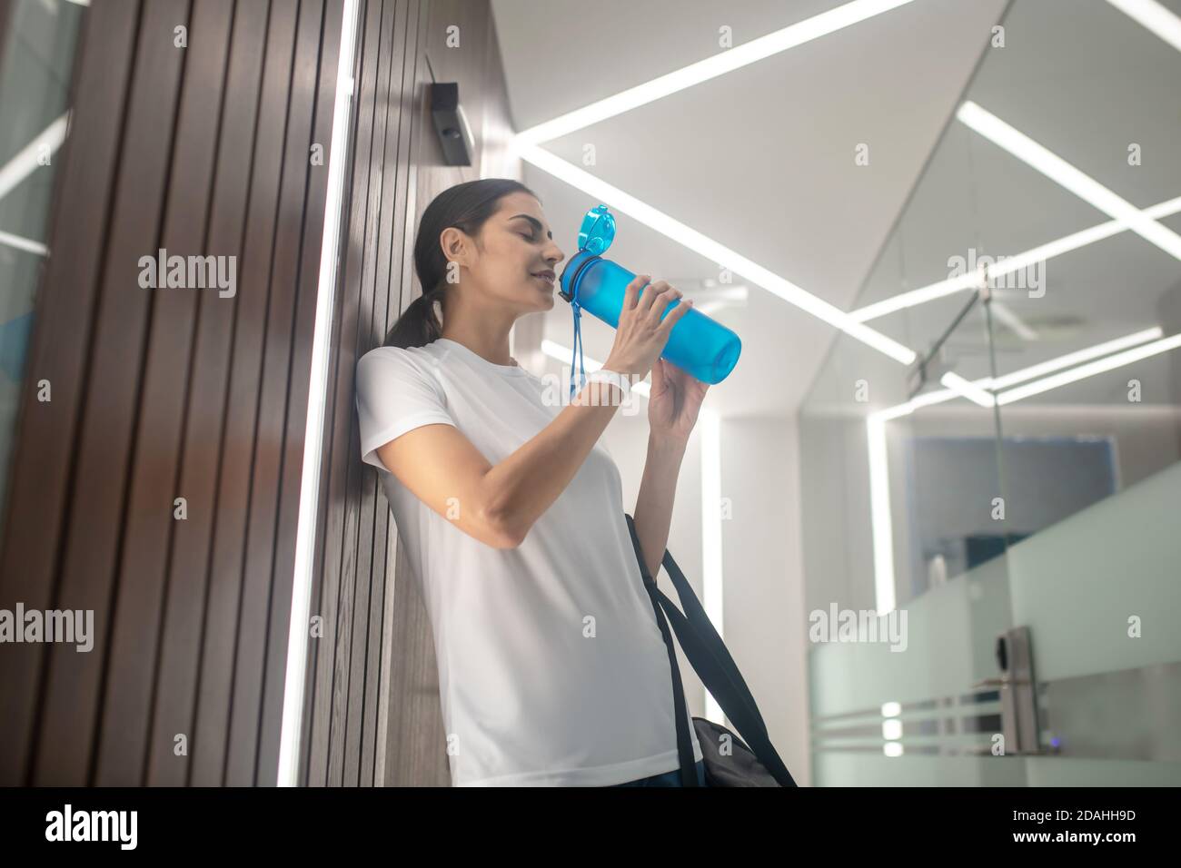 Dark-haired female with gym bag leaning on wall, drinking water Stock ...