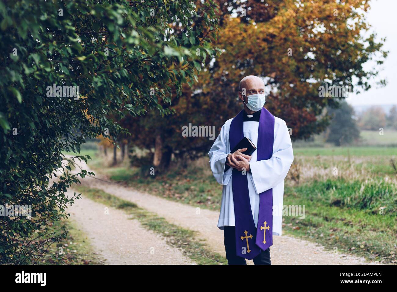 Catholic priest with a bible in a medical mask stands in a field in the ...