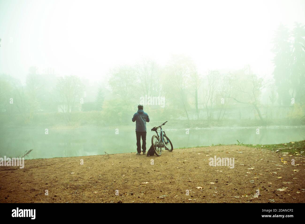 lone man standing near his bicycle in front of a lake in a foggy ...