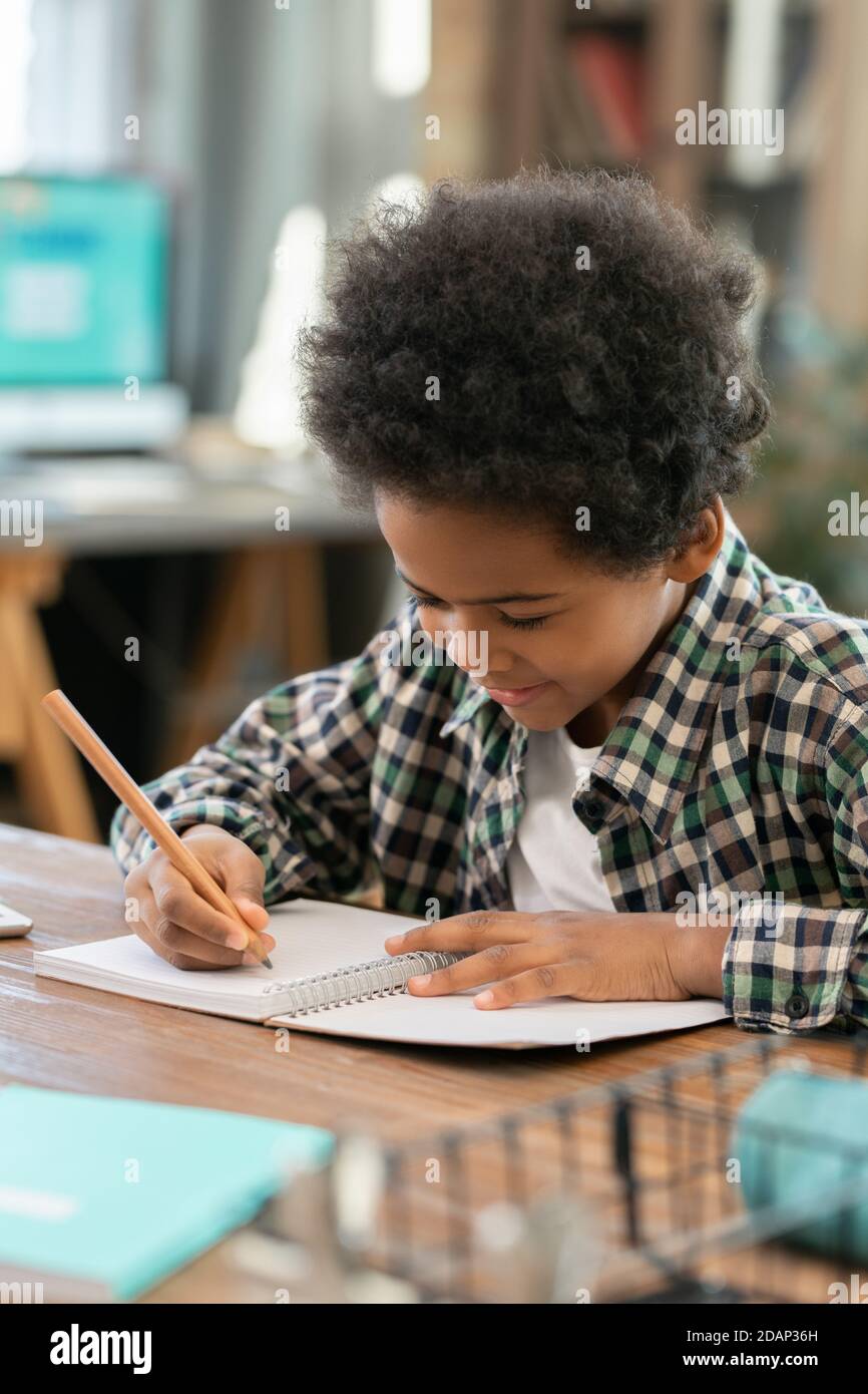 Cute elementary schoolboy of African ethnicity making notes in copybook ...