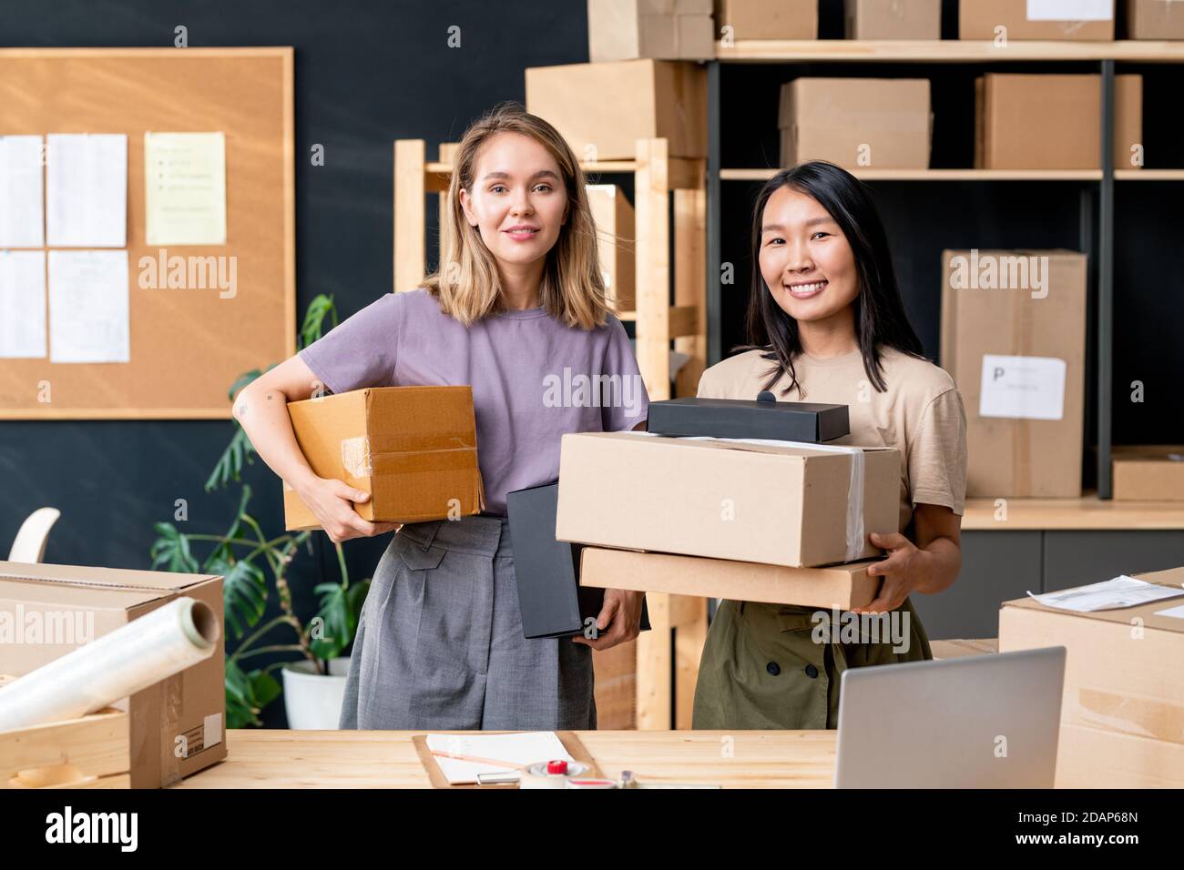 Two young female workers of online shop office holding packed boxes ...