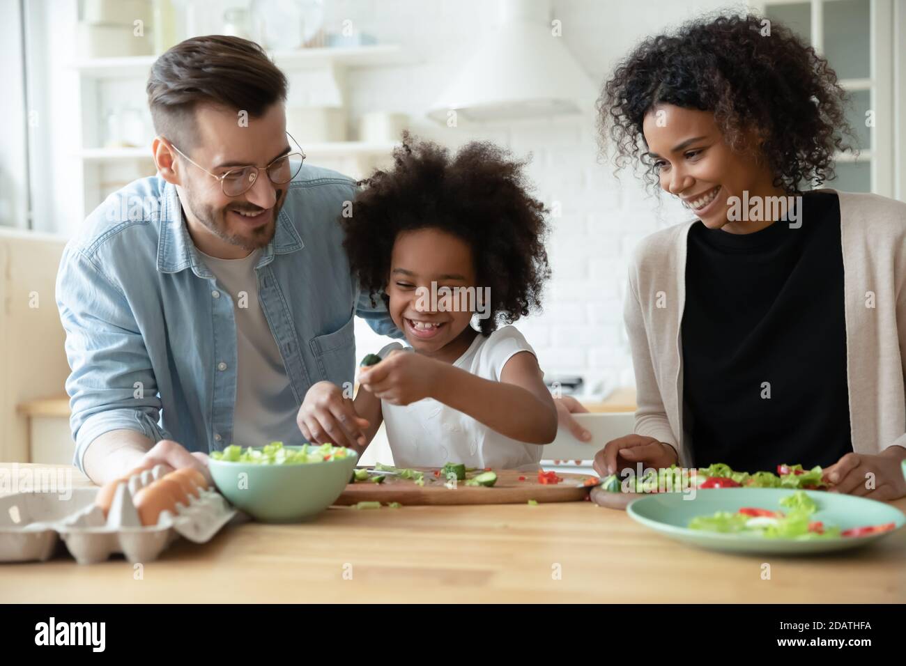 Happy diverse parents with little daughter cooking salad together Stock ...